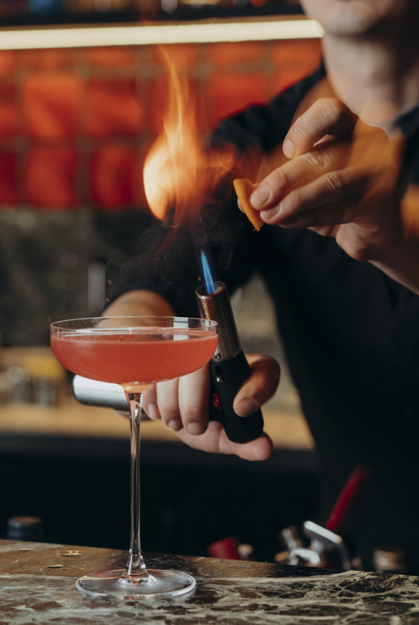 A bartender uses a lighter to create a flame above a pink cocktail on ...